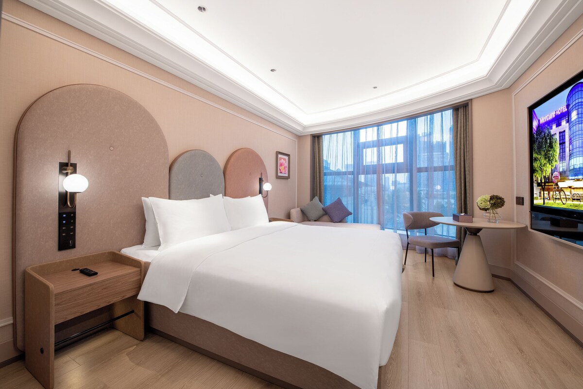 Quarto de hotel moderno com cama grande, mesa de cabeceira, televisão e vista para a cidade.