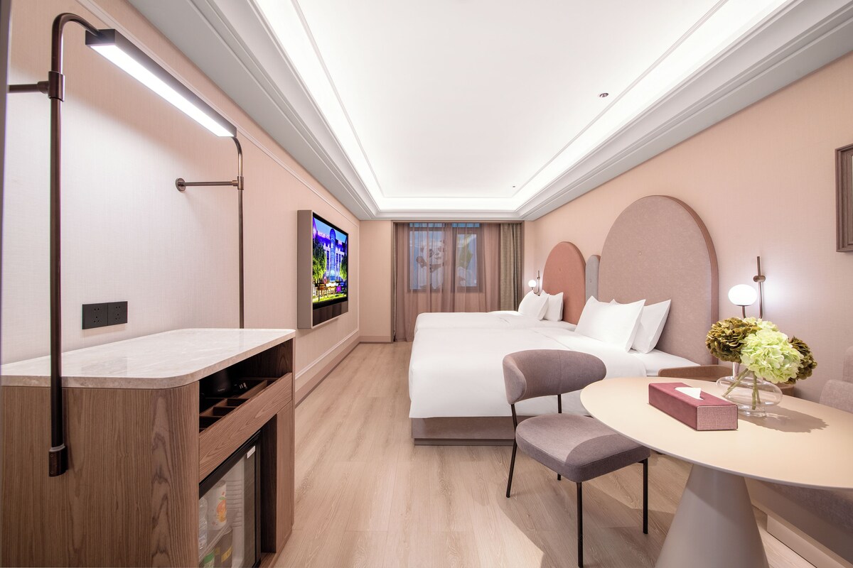 Quarto de hotel moderno com uma cama, uma escrivaninha, uma cadeira, uma televisão e uma mesinha com um vaso de flores.