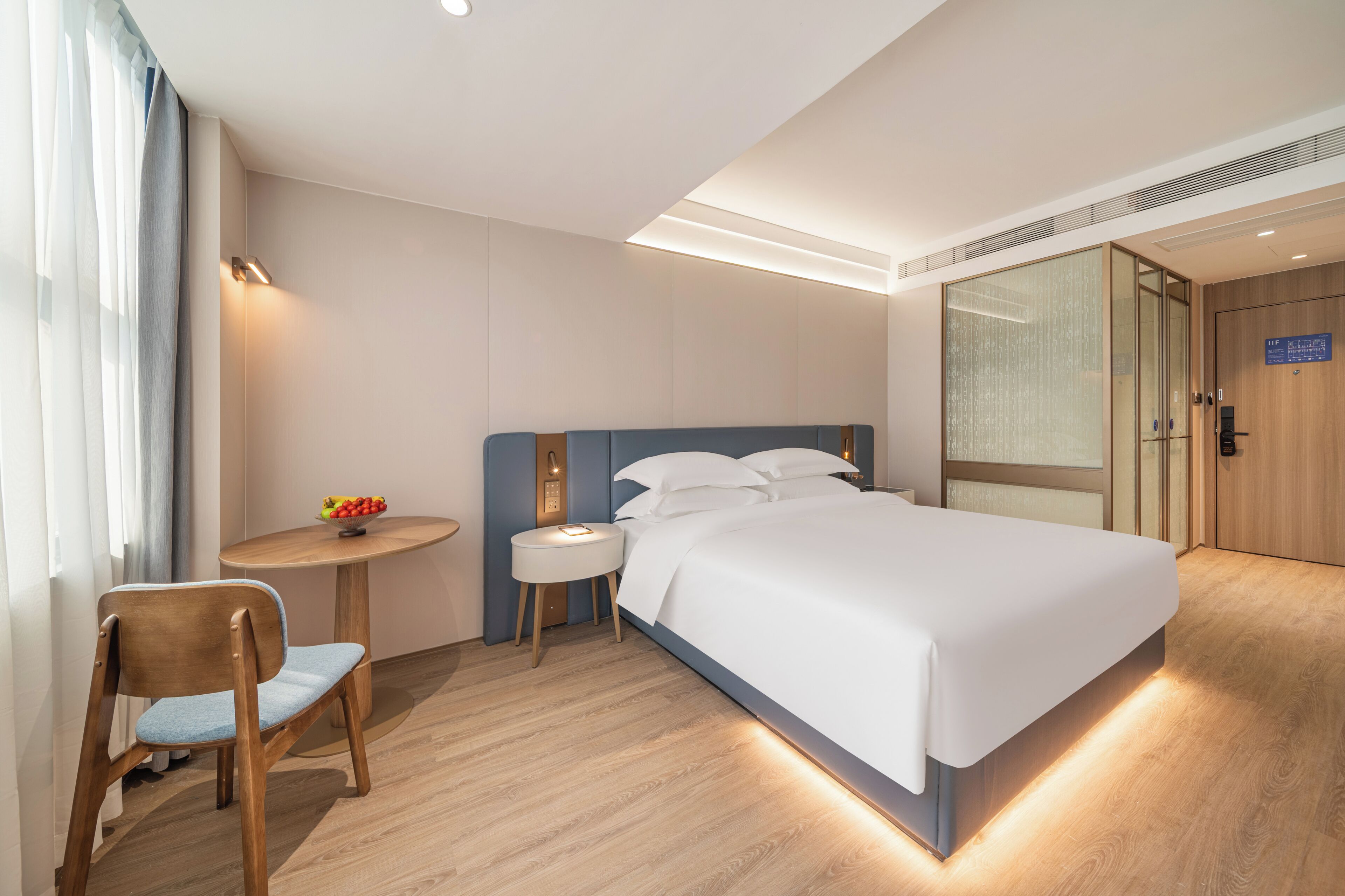 Quarto de hotel com uma cama, uma mesinha com frutas, uma cadeira e uma escrivaninha com uma luminária.