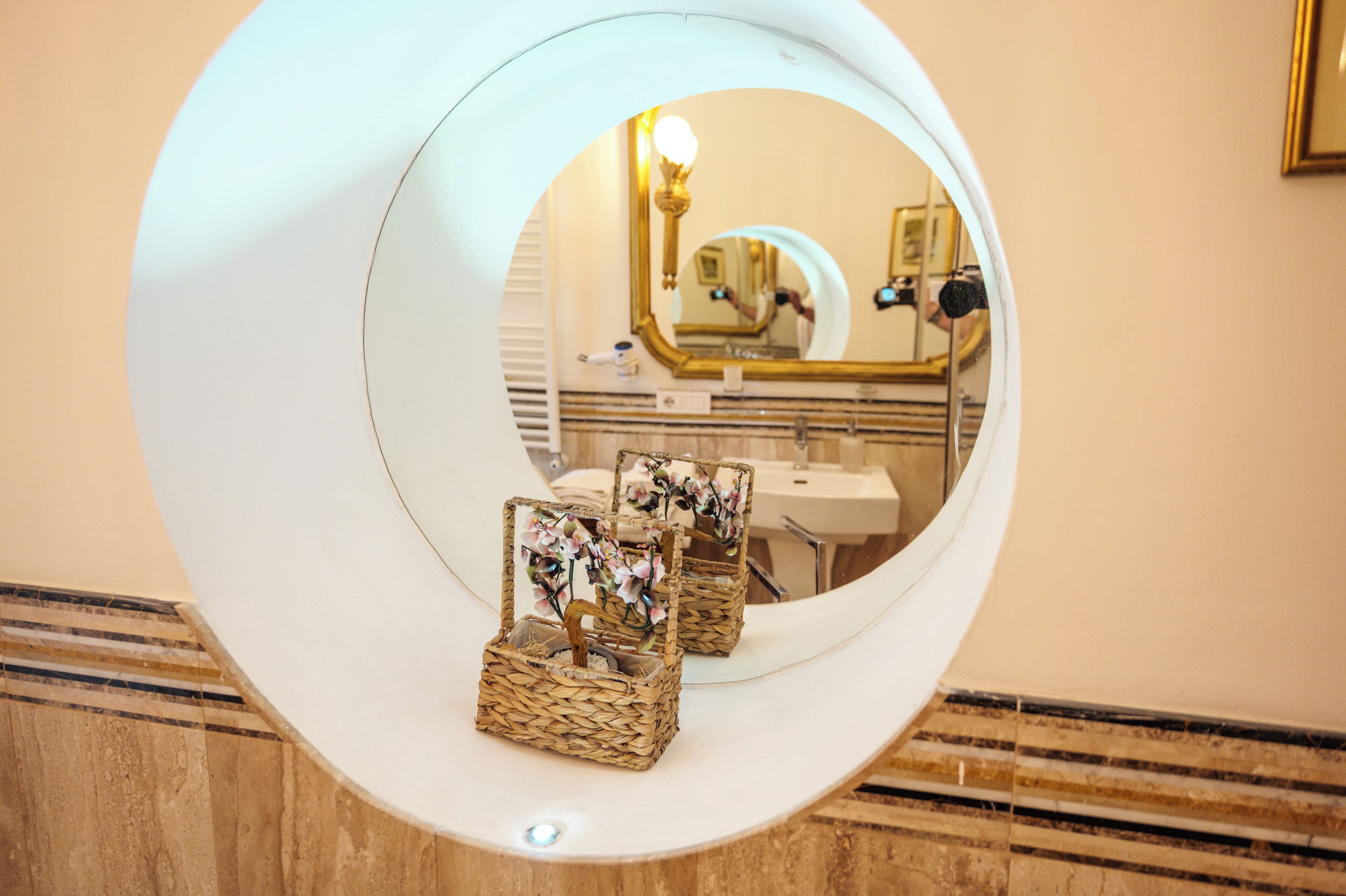 Espelho circular com moldura branca, refletindo o interior de uma casa de banho com lavatório, espelho e cestos decorativos.