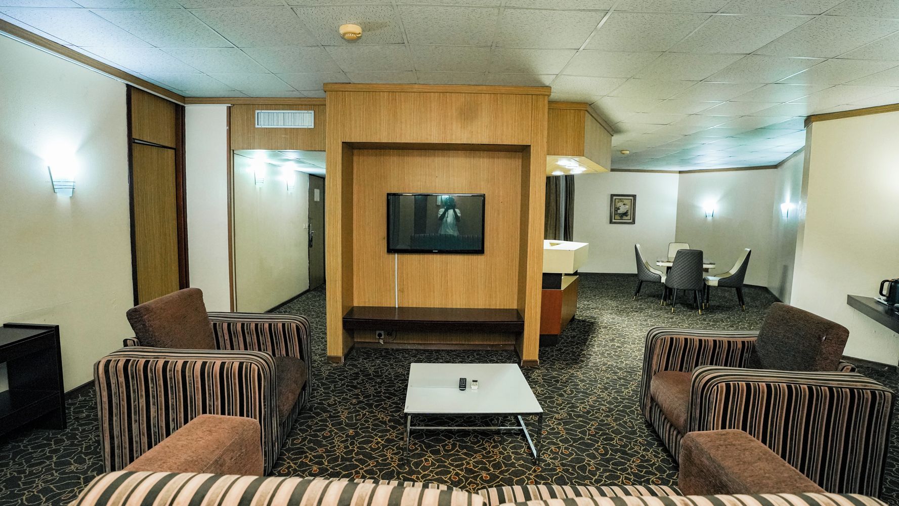 Quarto de hotel com televisão de tela plana fixada em um painel de madeira, poltronas listradas, uma pequena mesa branca e um espaço de refeições com cadeiras.