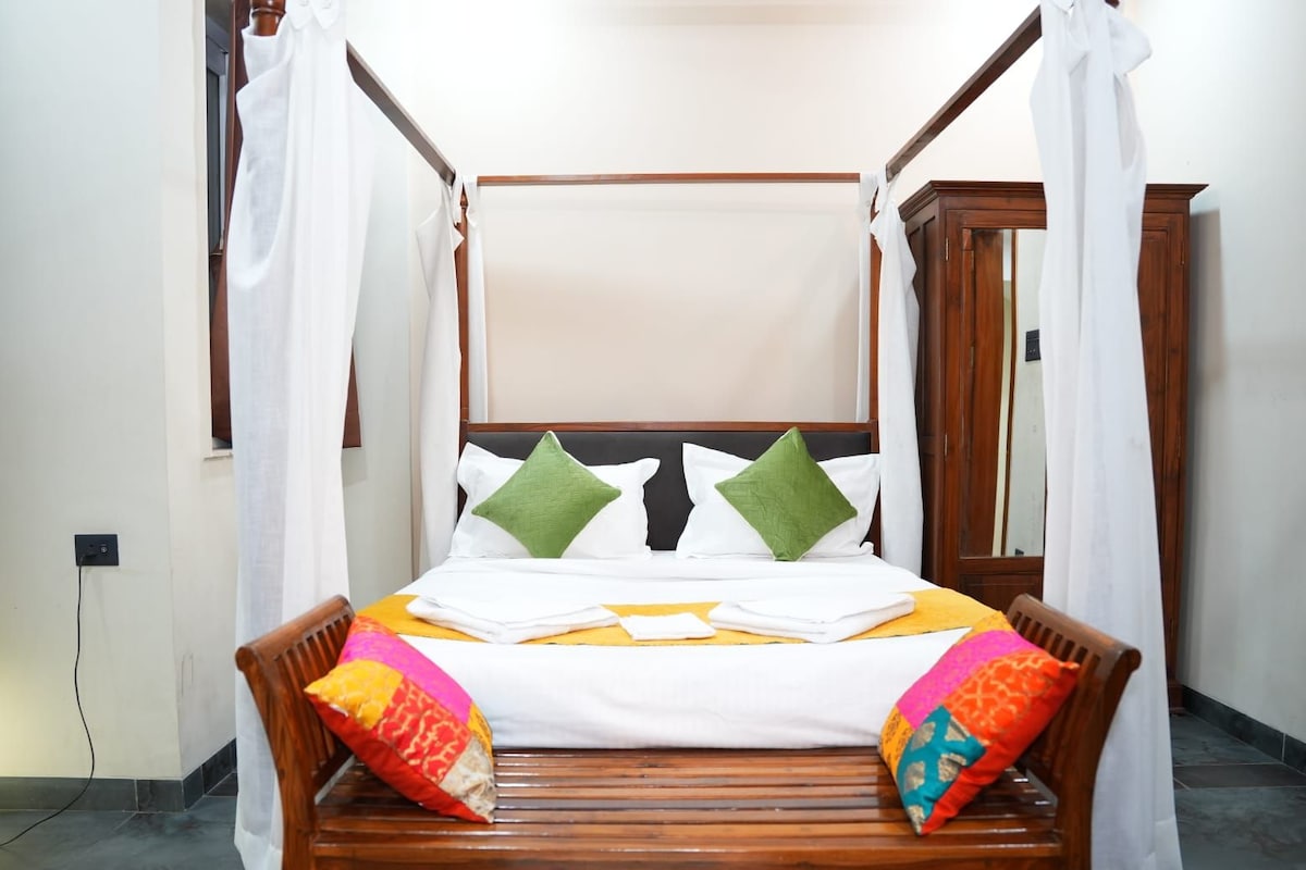 Uma cama com dossel, roupa de cama branca, almofadas verdes e colcha amarela, com cabeceira de madeira.