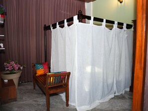 Um quarto com um banco de madeira, uma cama com dossel branca, um vaso de planta e uma prateleira fixada na parede.