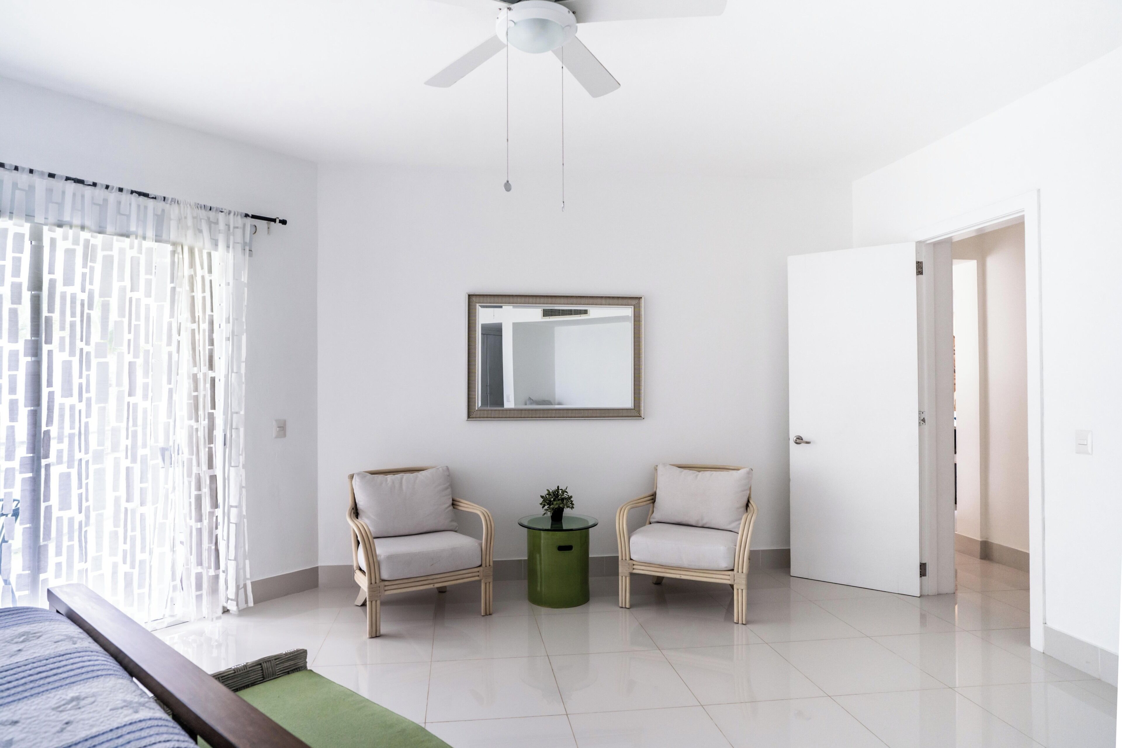 Quarto amplo com cama, duas poltronas, uma mesinha com uma planta, um ventilador de teto e uma porta.