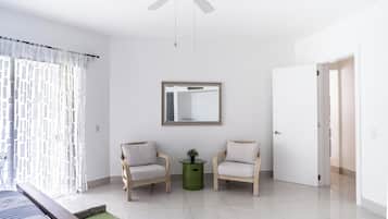 Quarto amplo com cama, duas poltronas, uma mesinha com uma planta, um ventilador de teto e uma porta.
