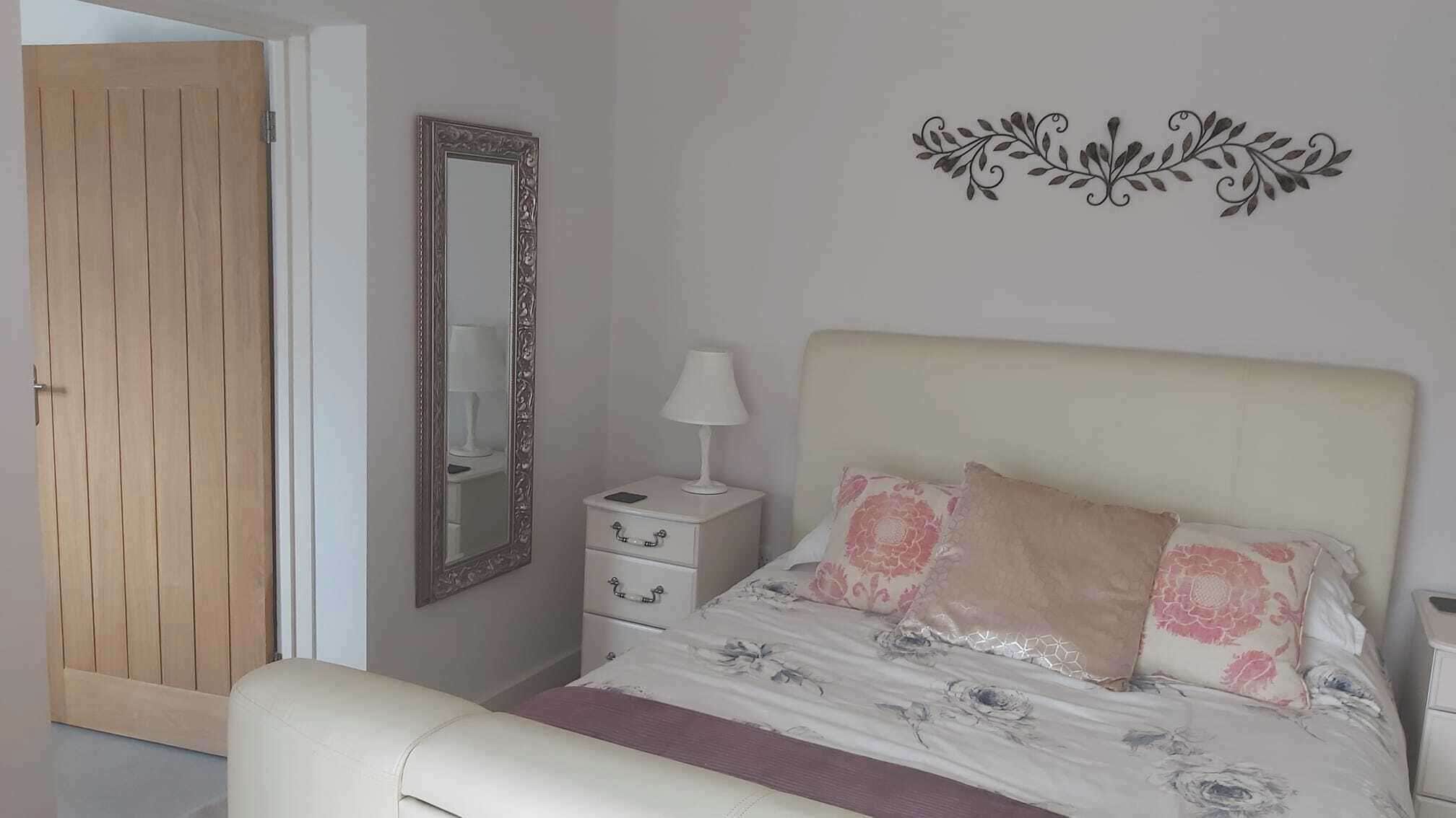 Quarto com guarda-roupa de madeira, mesa de cabeceira com abajur, cama com roupa de cama floral e um objeto decorativo na parede.