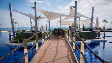 Um deck de madeira leva a uma área de piscina com espreguiçadeiras e guarda-sóis, com vista para o mar.