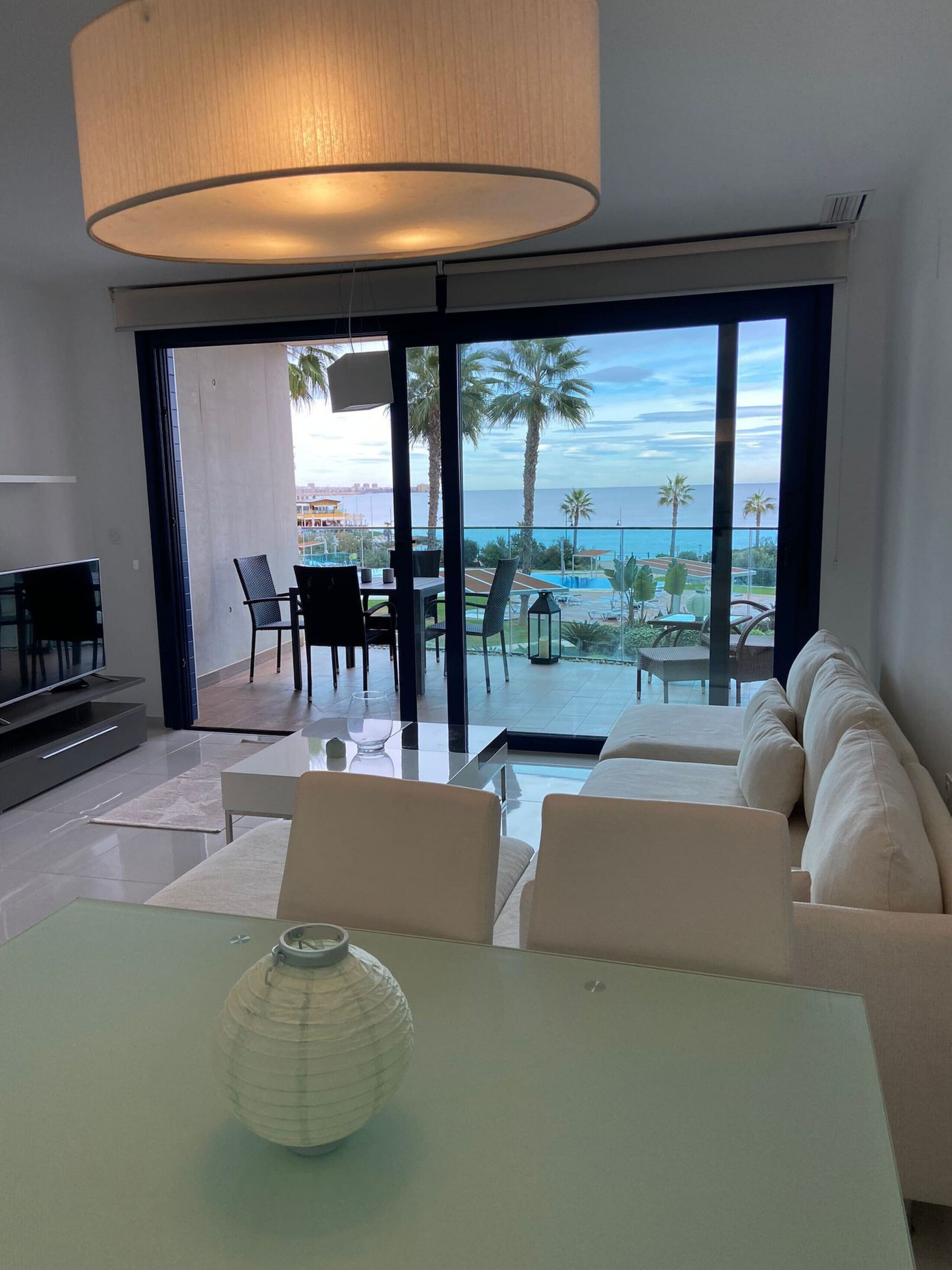 Sala de estar moderna com uma grande porta de vidro deslizante que dá para uma varanda com vista para o mar e palmeiras.