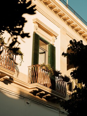 Um edifício com varanda, uma janela com persianas verdes e algumas plantas.