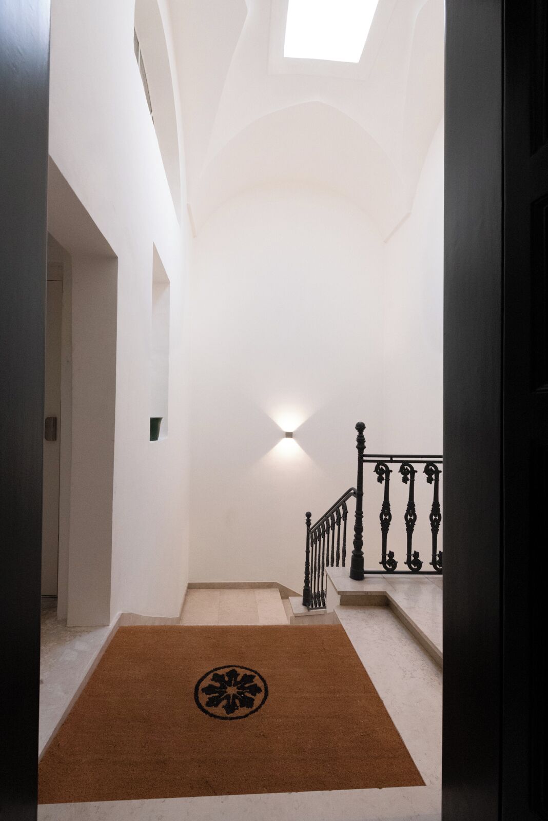 Uma escadaria com corrimão, um luminário na parede e um tapete de entrada marrom com um desenho circular.