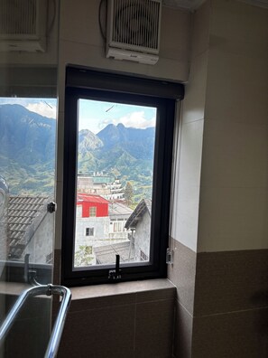 Casa de banho com janela que oferece vista para uma paisagem montanhosa e um edifício com telhado vermelho.