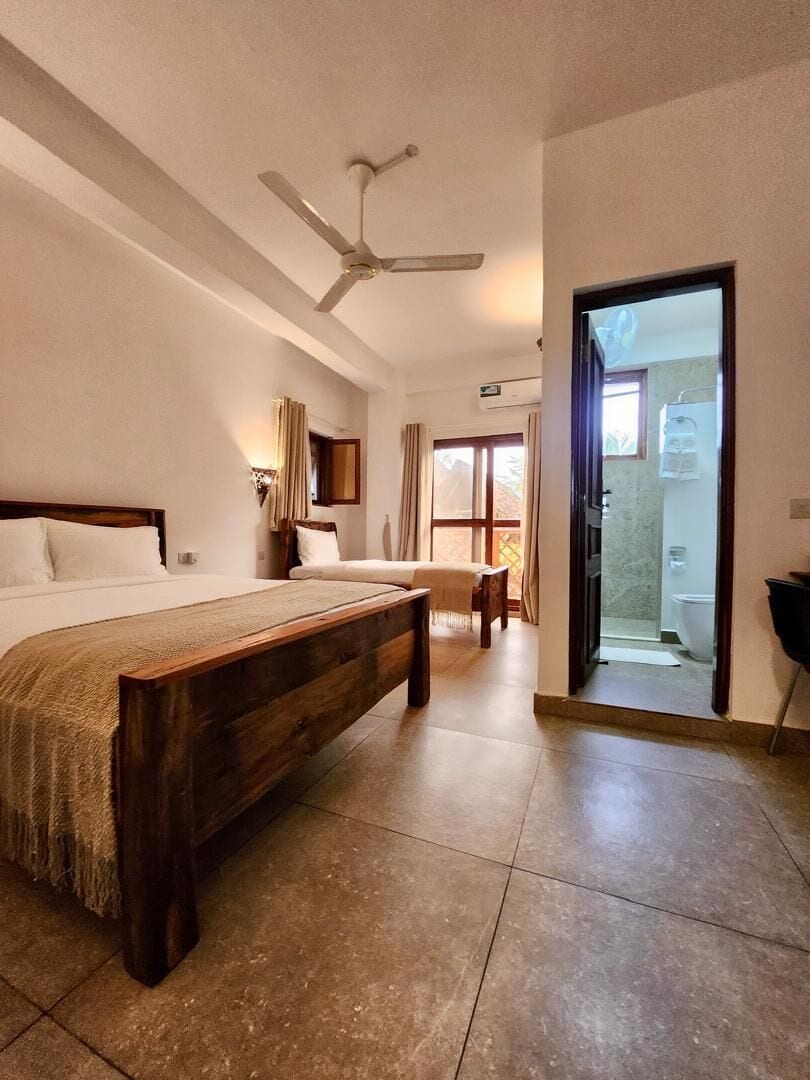 Quarto de hotel com uma cama grande, um banquinho de madeira, um ventilador de teto e vista para o exterior.