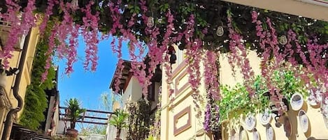 Uma área externa com mesas e cadeiras, decorações de flores penduradas e fachada de prédio com a placa 'Bellis Siciliani Ristorante Autentico Siciliano'.