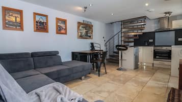 Uma sala moderna com um sofá cinzento, uma área de jantar com mesa e cadeiras, uma cozinha com eletrodomésticos, e uma escada.