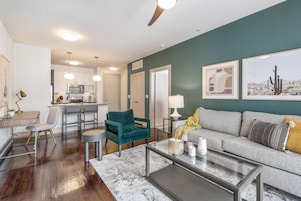 Uma sala de estar moderna com um sofá cinza, uma poltrona azul-turquesa e uma mesa de centro de vidro. H á uma área de cozinha com armários brancos e um conjunto de jantar.