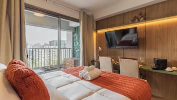 Quarto de hotel com uma cama grande, uma televisão presa à parede, uma varanda com vista para edifícios e uma máquina de café em uma mesinha.