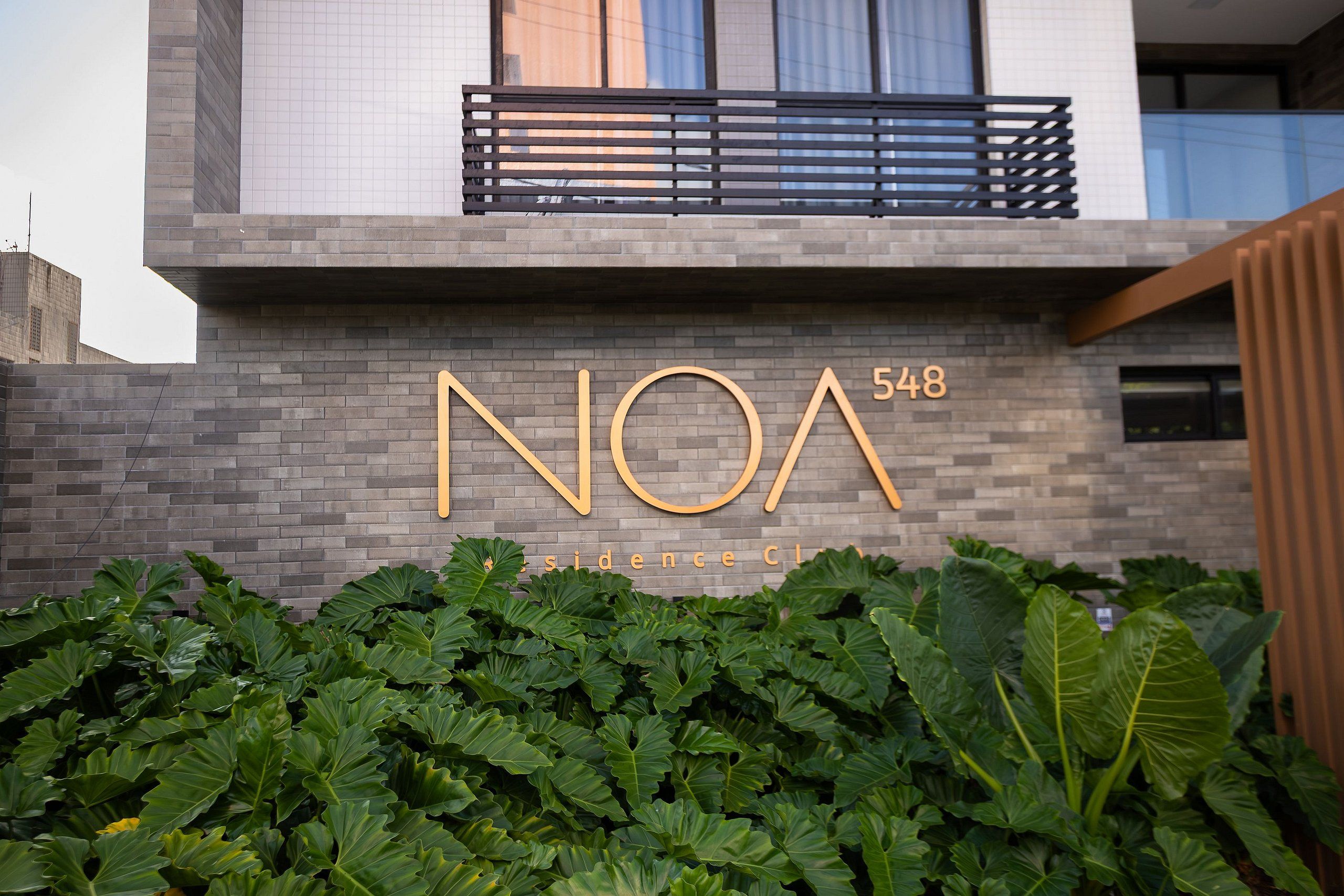 Um edifício com uma grande placa indicando 'NOA 548 Residence Club' acima de uma parede de tijolos.