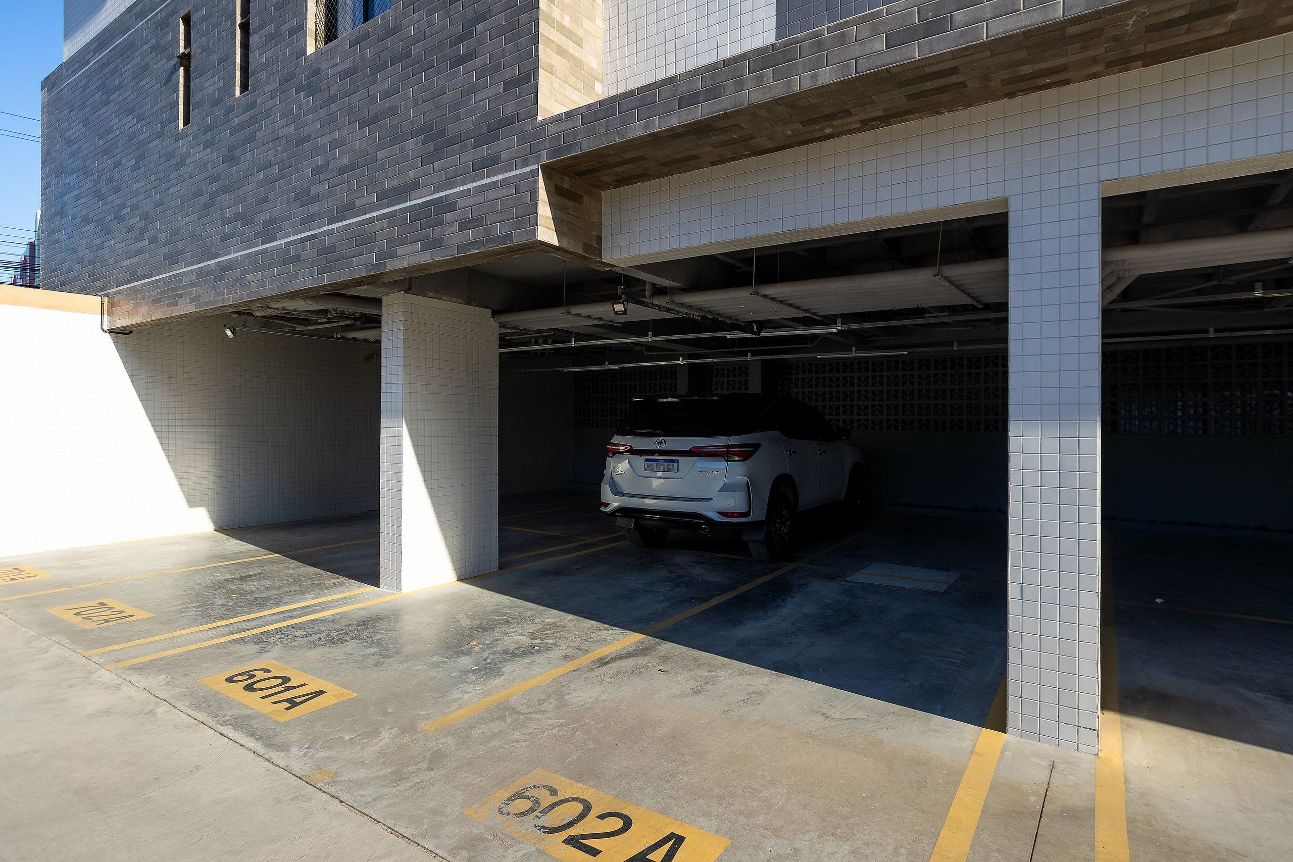 Um estacionamento com vagas demarcadas, um carro branco estacionado e um edifício com paredes e colunas revestidas de azulejos.