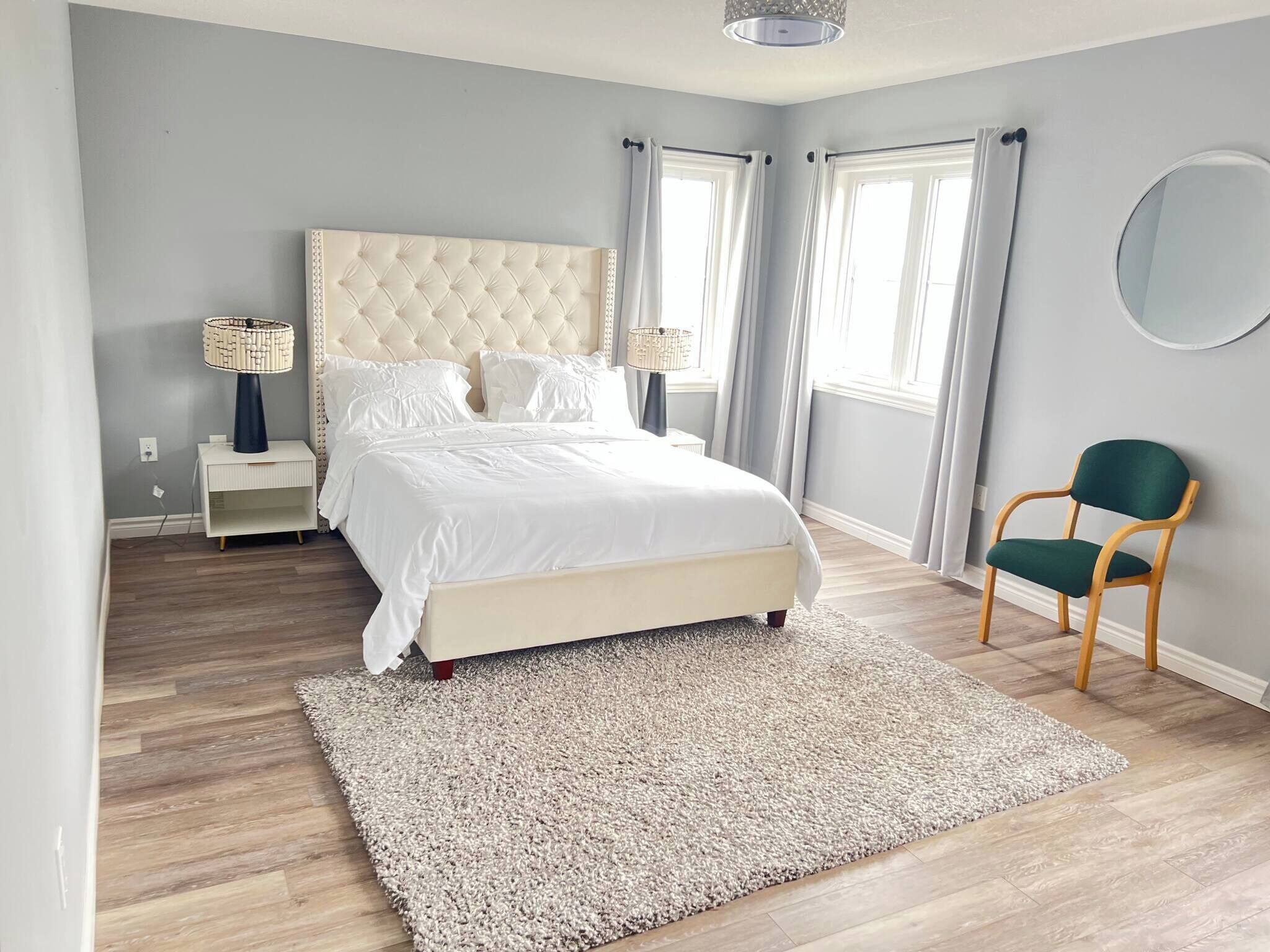 Quarto com cama de cabeceira estofada, duas luminárias de apoio, uma poltrona verde e um espelho grande na parede.