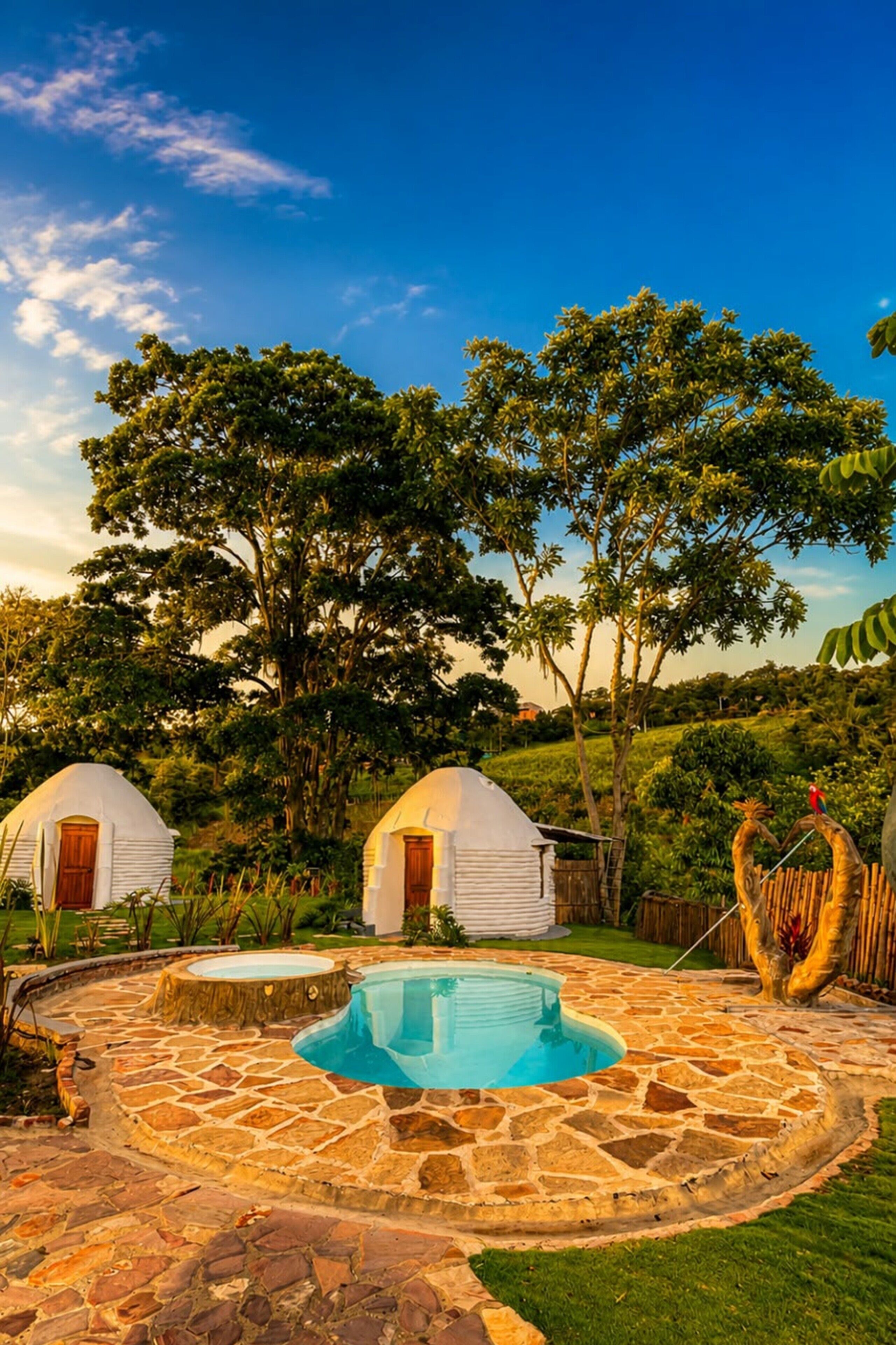 Uma área de piscina com piso de pedra, rodeada por duas yurts em formato de cúpula e uma grande árvore.