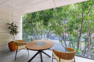 Quarto moderno com uma mesa redonda e duas cadeiras, uma planta em vaso e uma janela grande com vista para árvores e edifícios.