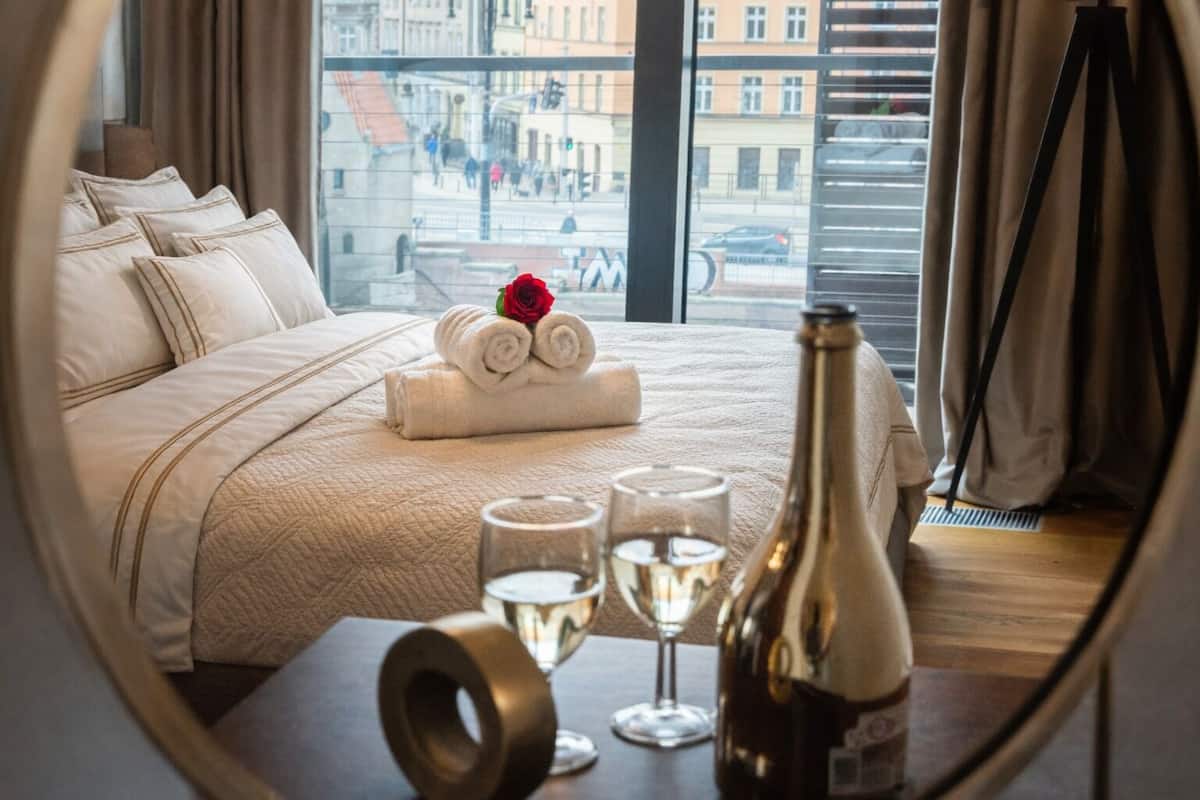 Quarto de hotel com cama, janela com vista para a cidade, mesa com garrafa e copos, e espelho refletindo o quarto.