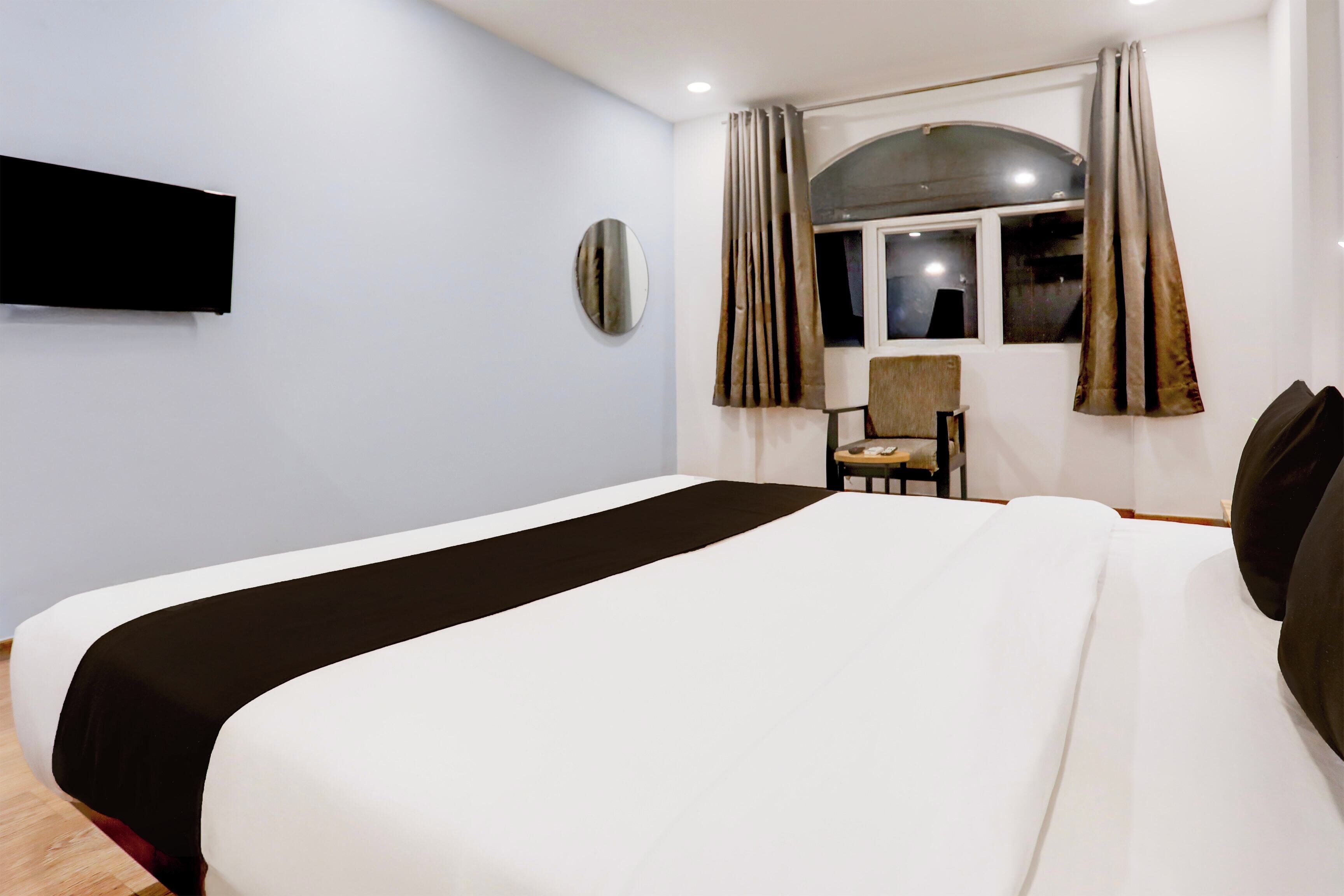 Quarto de hotel com cama, cadeira, televisão fixada na parede e janela com cortinas.