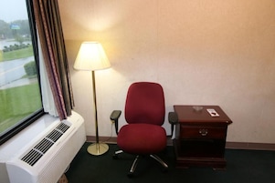 Quarto de hotel com janela, uma cadeira de escritório vermelha, luminária de chão e mesa de cabeceira de madeira.