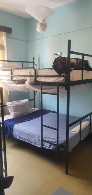 Quarto com beliche, ventoinha no teto, janela com cortinas e uma mochila em cima da cama.