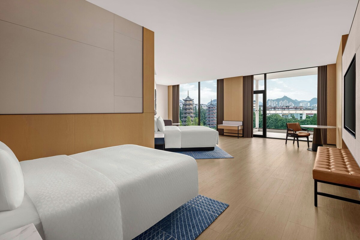 Quarto de hotel com cama, área de estar com mesa e cadeiras, e janelas amplas com vista para a cidade.