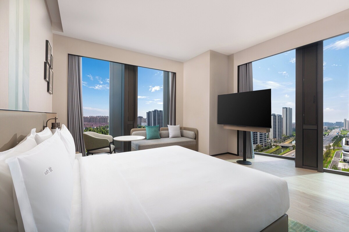 Quarto de hotel com uma cama grande, televisão, sofá e vista para a cidade.
