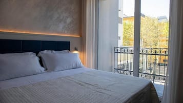 Quarto de hotel com cama grande, cabeceira, janela com vista para a rua e varanda com grade de ferro forjado.
