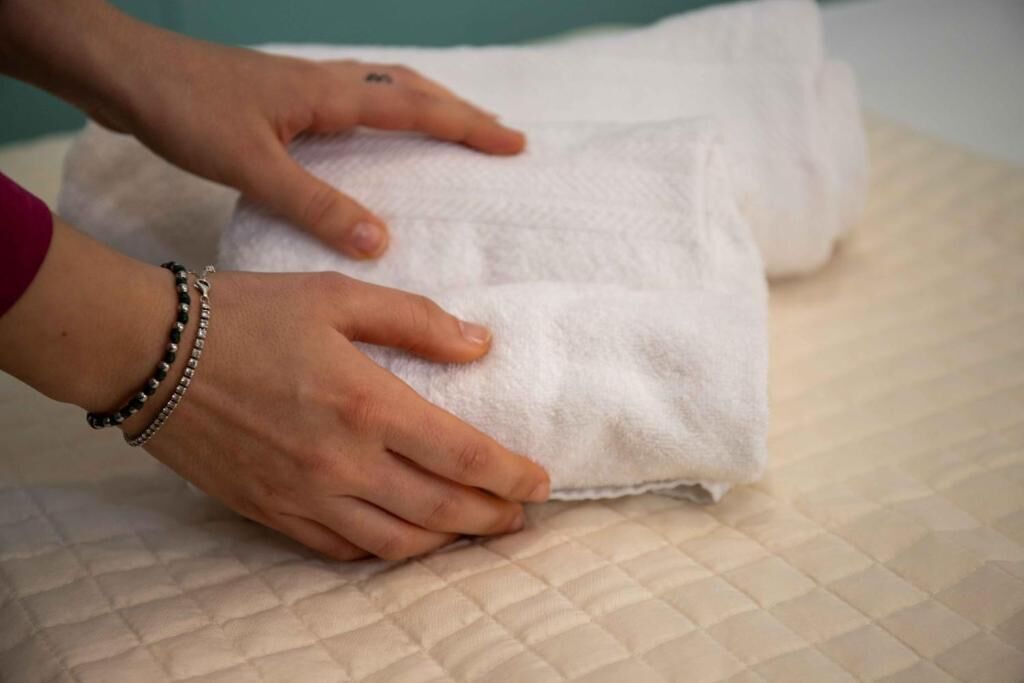 Mãos dobrando uma toalha branca sobre uma cama com cobertura acolchoada.