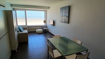 Quarto compacto com mesa de jantar, cadeiras, sofá, radiador e uma grande janela com vista para a praia.