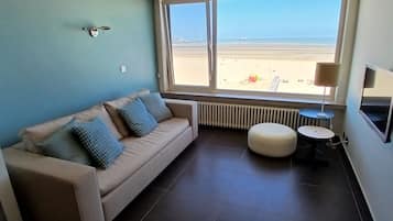 Quarto com sofá bege, uma pequena mesa redonda, televisão e vista para a praia.