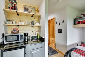 Uma cozinha com micro-ondas, cafeteira e armários, e um quarto com beliche e prateleira na parede.