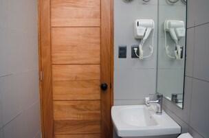 Casa de banho com porta de madeira, pia branca, espelho e dois secadores de cabelo fixados na parede.