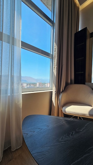 Quarto com cama, poltrona, janela com cortinas e vista para edifícios e montanhas.
