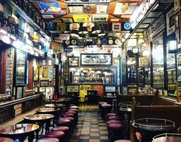 Um bar antigo com chão xadrez, vários bancos e um balcão com diversos objetos.