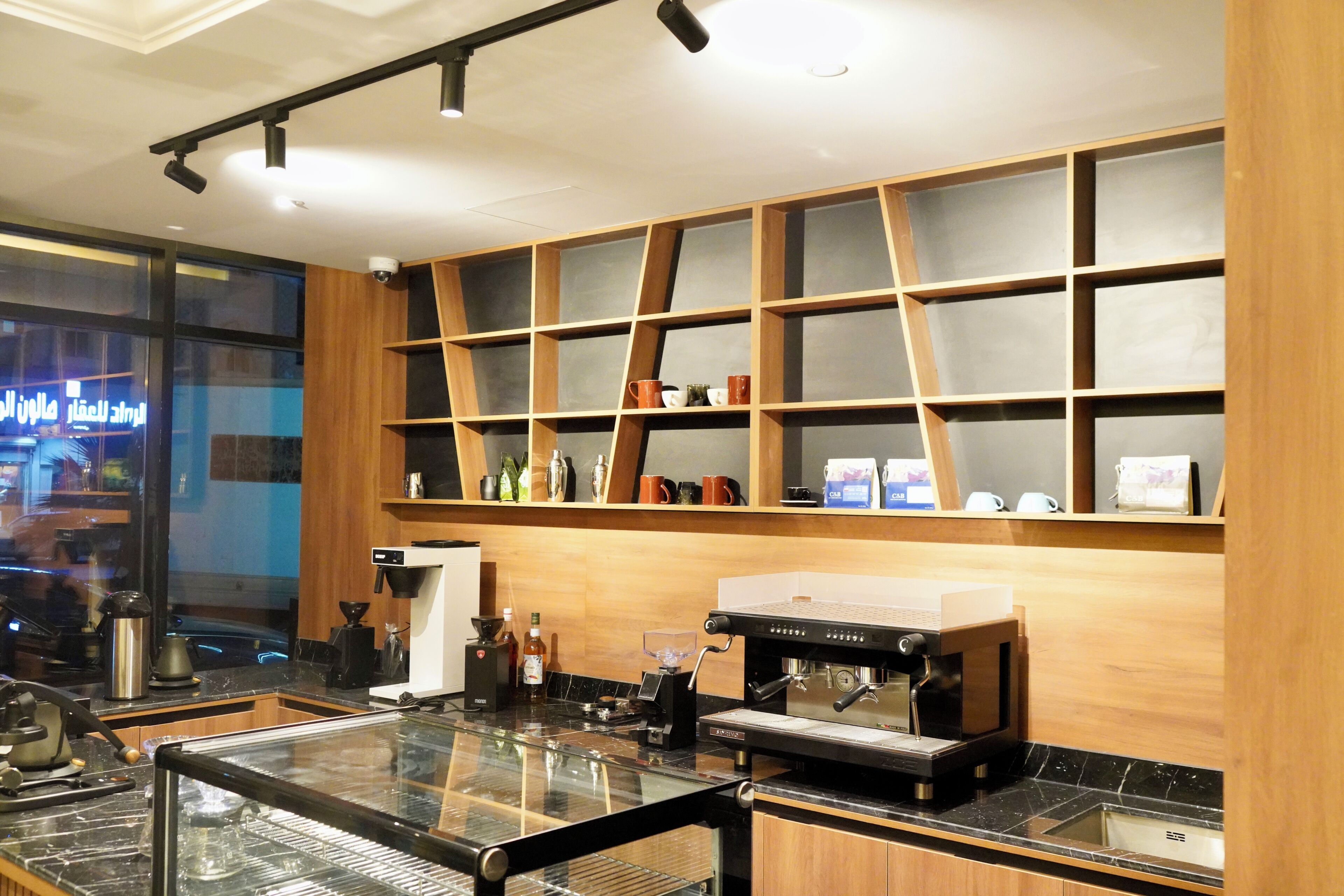 Interior de uma cafeteria moderna com um balcão de vidro, máquina de café expresso e prateleiras de madeira com diversos itens.