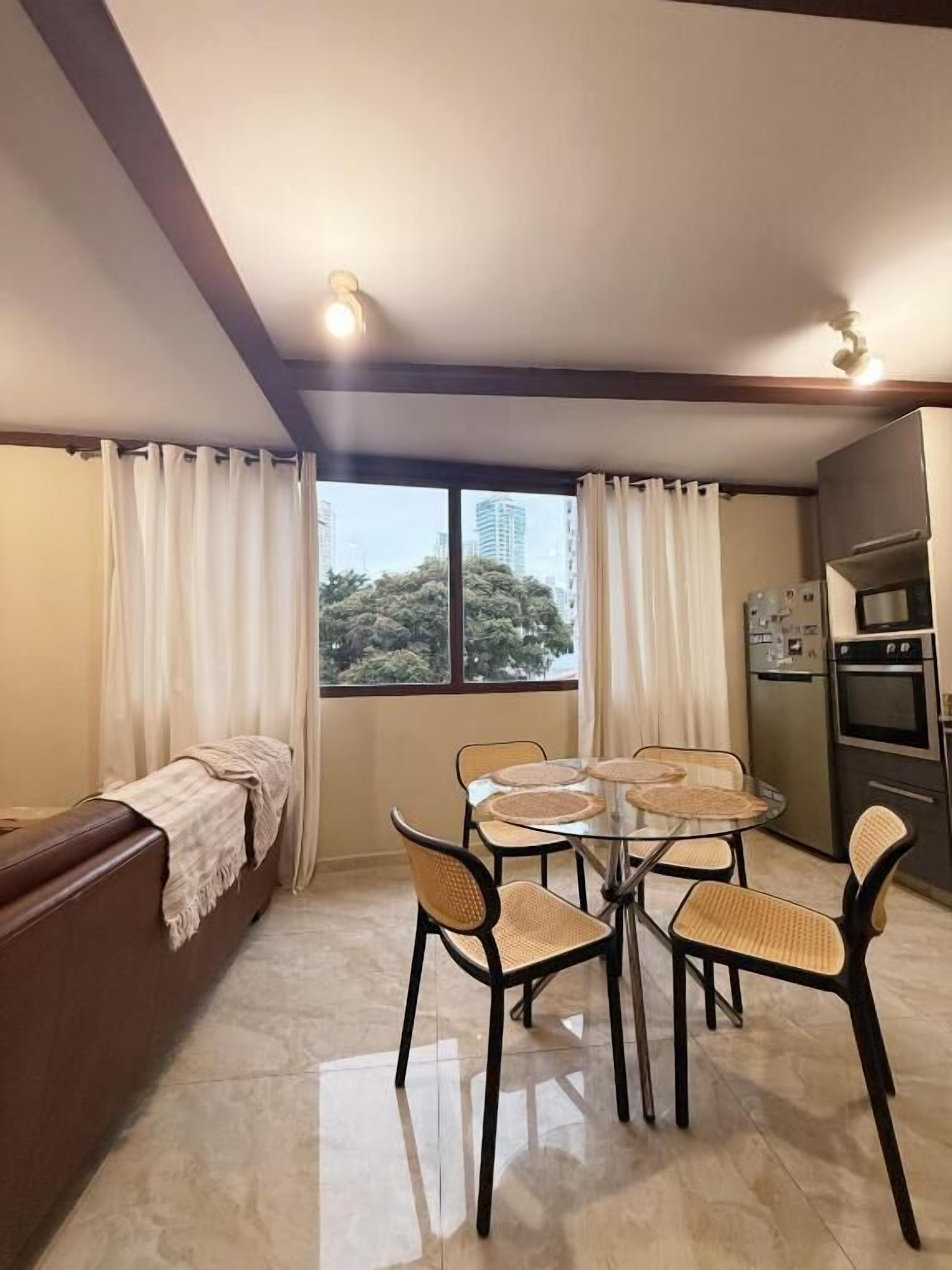 Uma sala de jantar com uma mesa redonda e quatro cadeiras, um sofá e uma cozinha equipada.