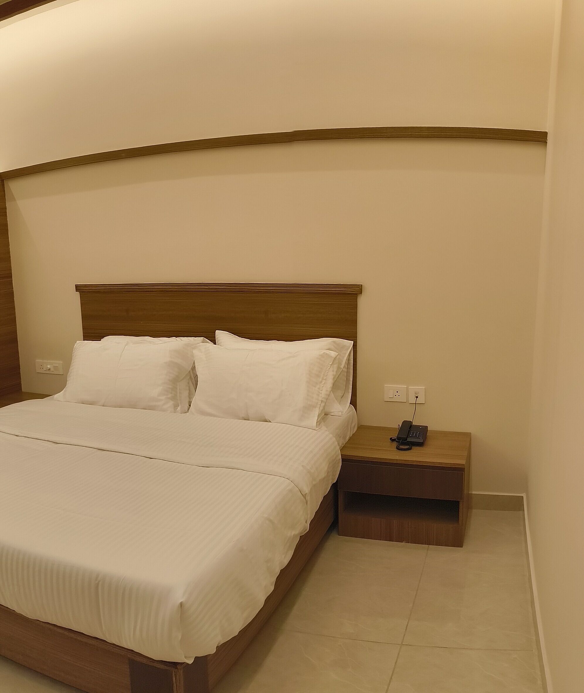 Uma cama bem arrumada com lençóis brancos, cabeceira de madeira, mesa de cabeceira com telefone e tomada na parede.