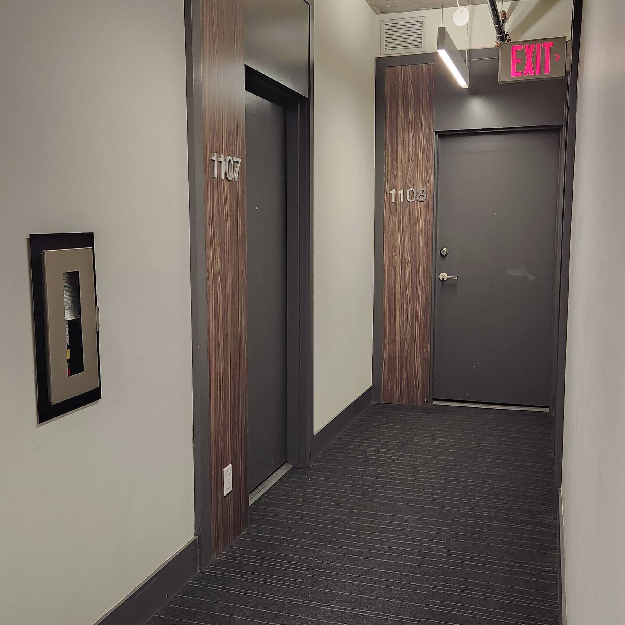 Um corredor com duas portas, uma com a placa 1107 e outra com a placa 1108, e um sinal de SAÍDA acima da porta à direita.