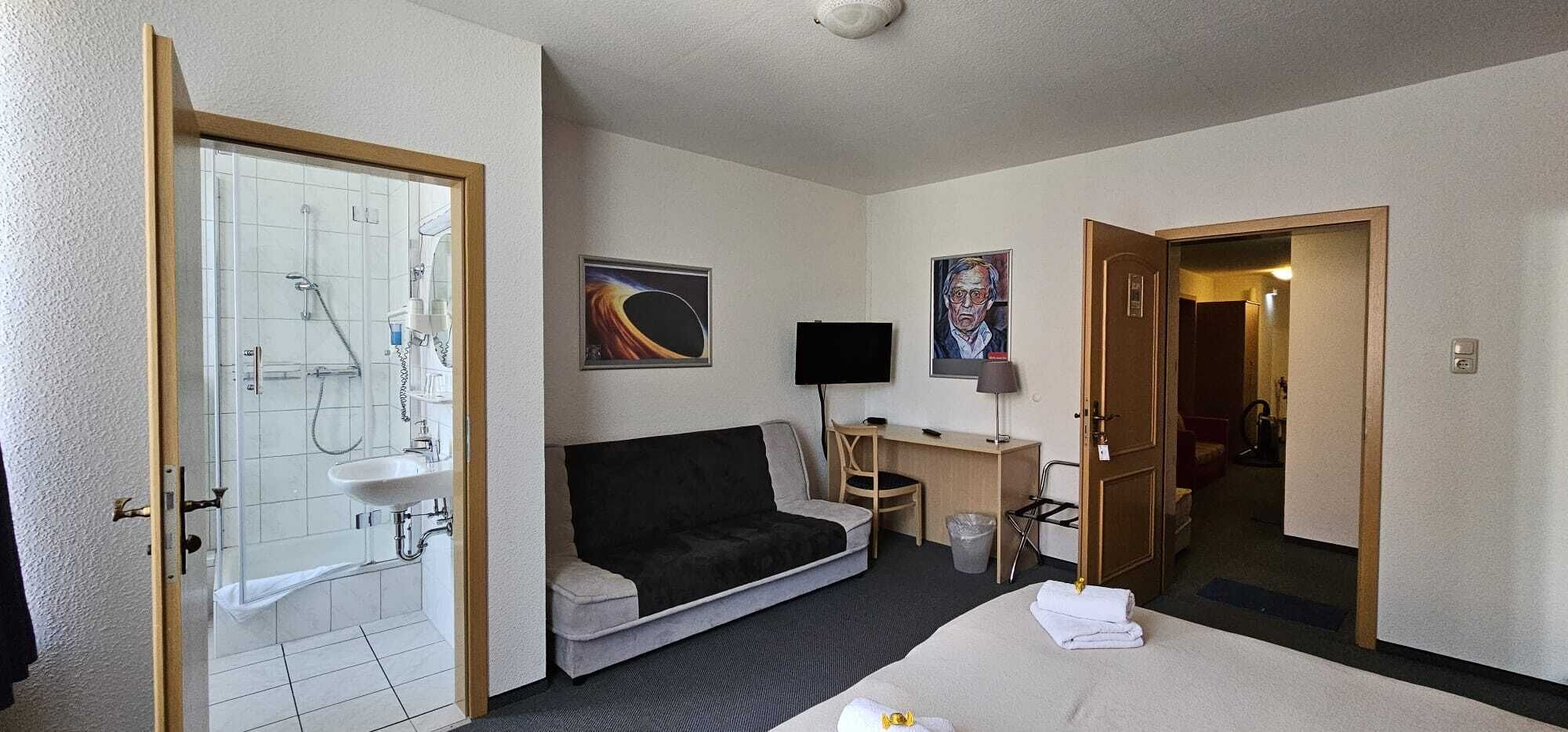 Quarto de hotel com cama, escrivaninha, cadeira, sofá, televisão e banheiro com chuveiro e pia.