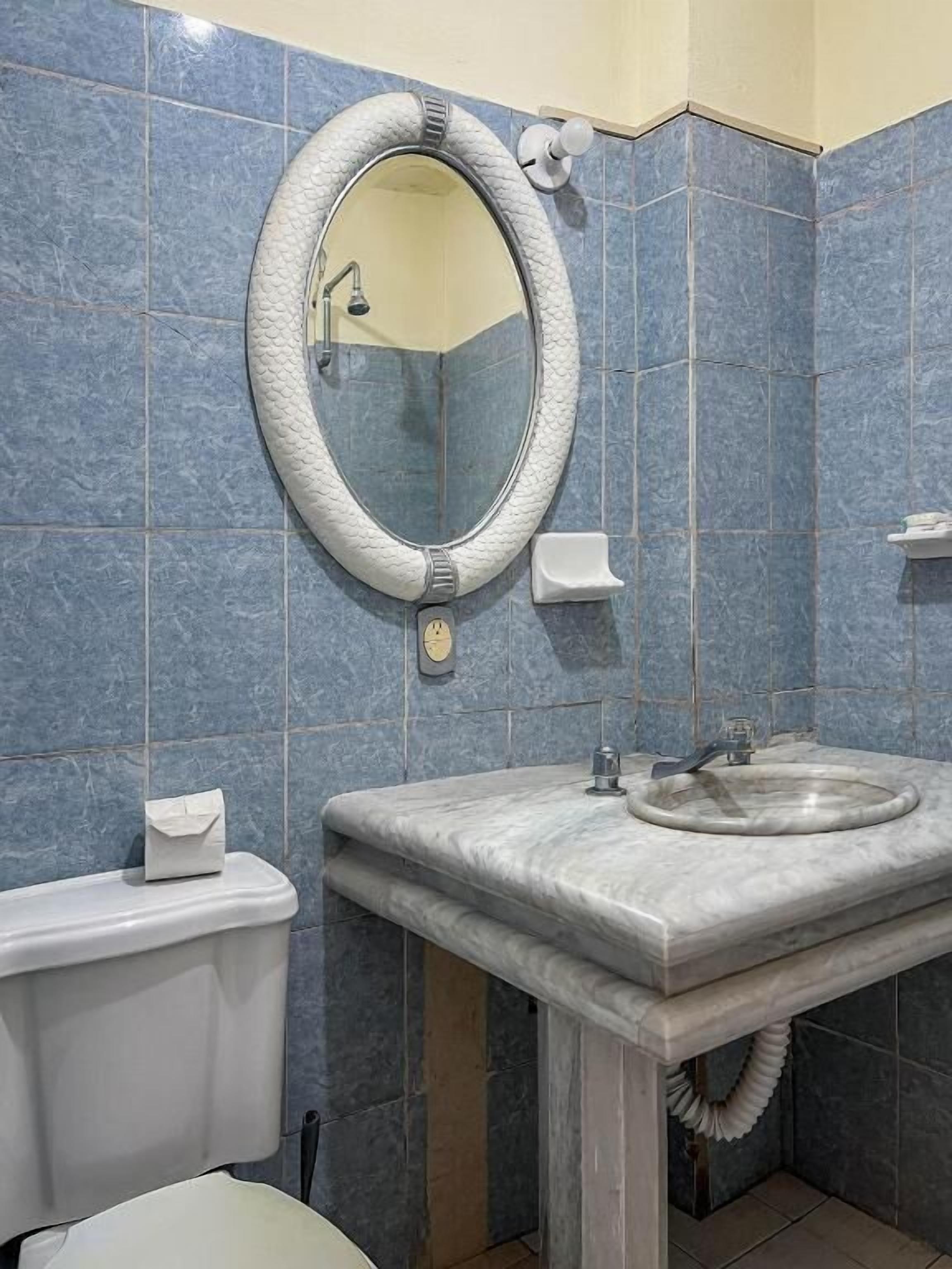Casa de banho com lavatório de mármore, espelho redondo com moldura texturizada, sanita e dispensador de sabonete.