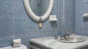 Casa de banho com lavatório de mármore, espelho redondo com moldura texturizada, sanita e dispensador de sabonete.