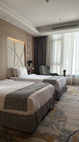 Quarto de hotel com duas camas, cabeceira com design geométrico, mesa pequena com abajur e uma janela grande com cortinas leves.