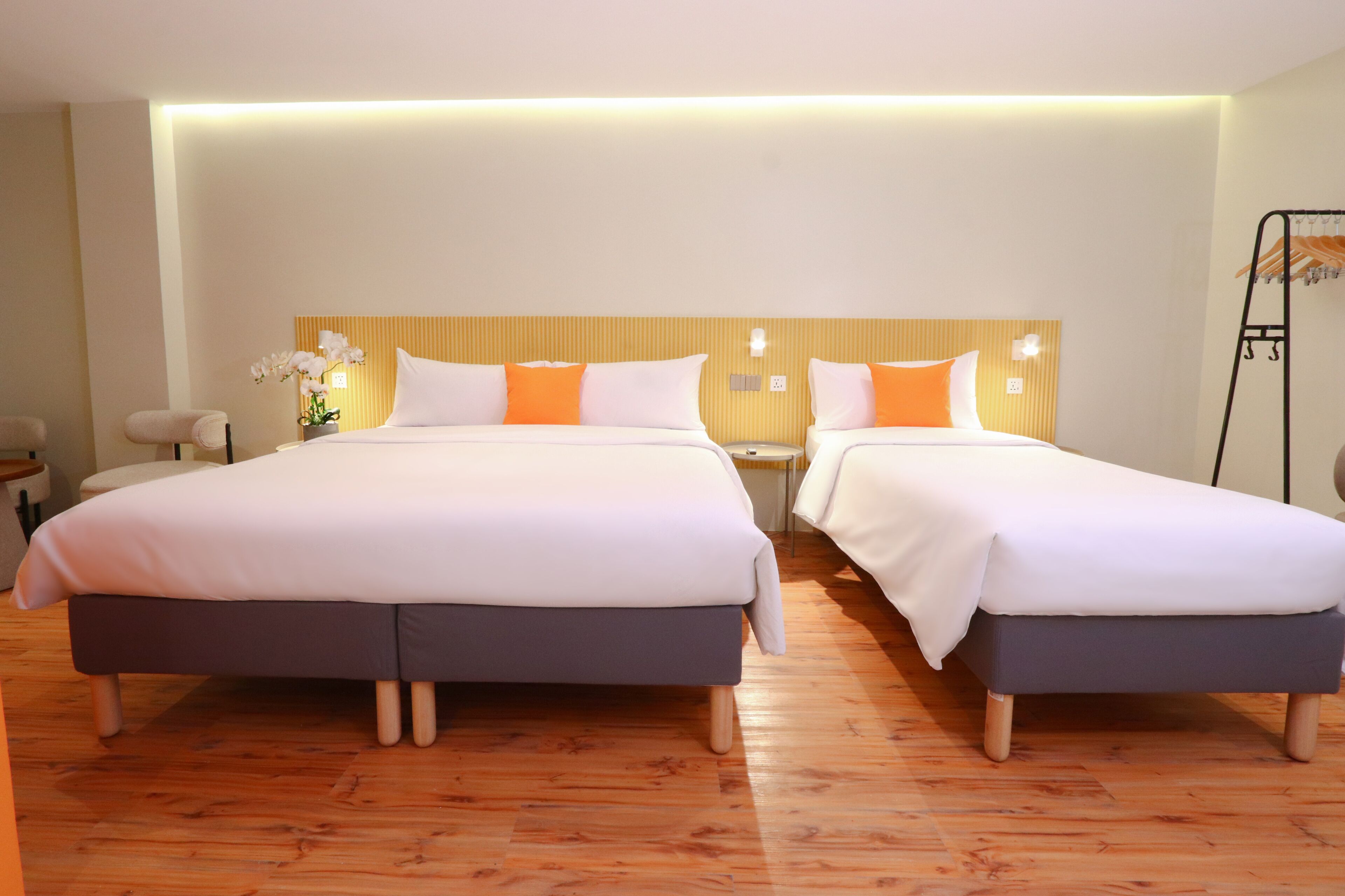 Quarto de hotel com duas camas, piso de madeira, luminária fixada na parede e uma mesinha com um vaso de flores.