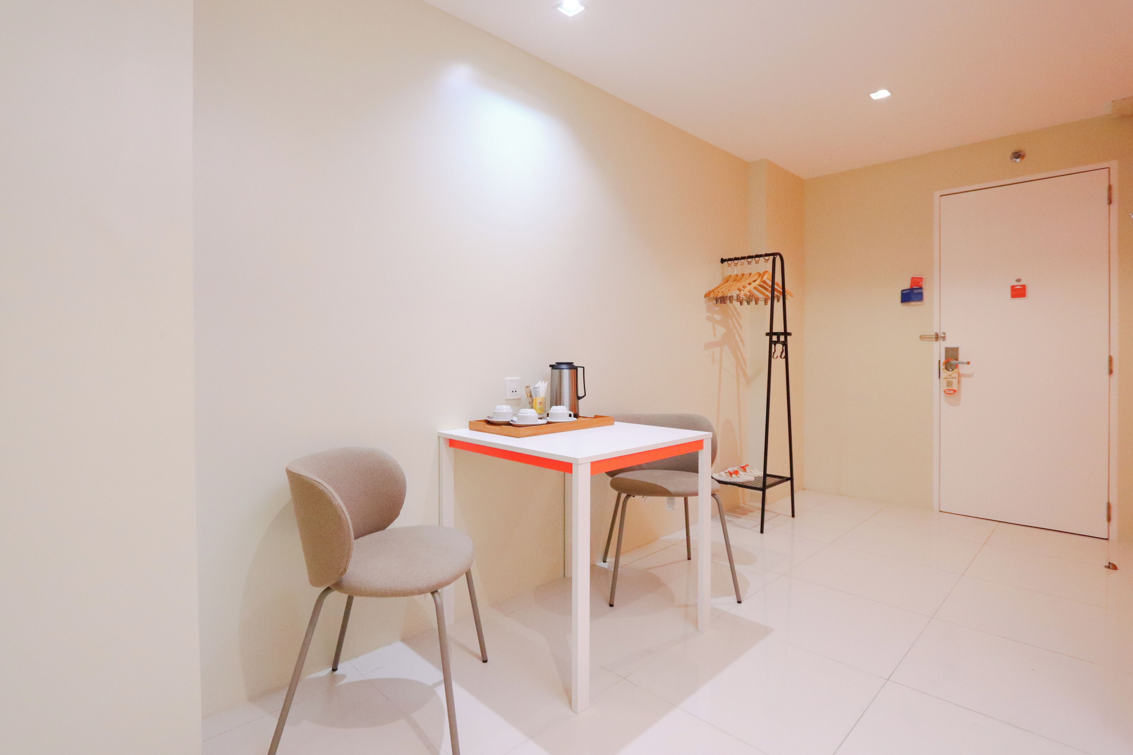 Quarto de hotel com uma mesa branca, duas cadeiras, um cabideiro e uma porta.