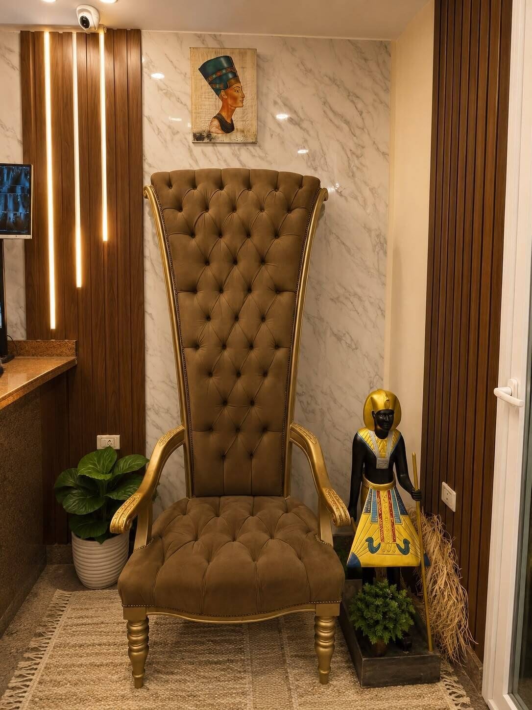 Uma poltrona marrom com textura, detalhes dourados, uma figura decorativa egípcia e uma parede moderna com padrão de mármore.
